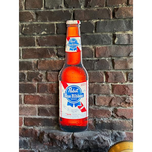 思わず「冷えてる!?」って聞きたくなるほどリアル！こちらはアメリカの老舗ビールブランドパブスト・ブルーリボンのボトルをモチーフにしたブリキ看板です。光沢のあるブリキ素材に ビール瓶特有の琥珀色がしっかり再現されていてさらに表面には水滴まで描...