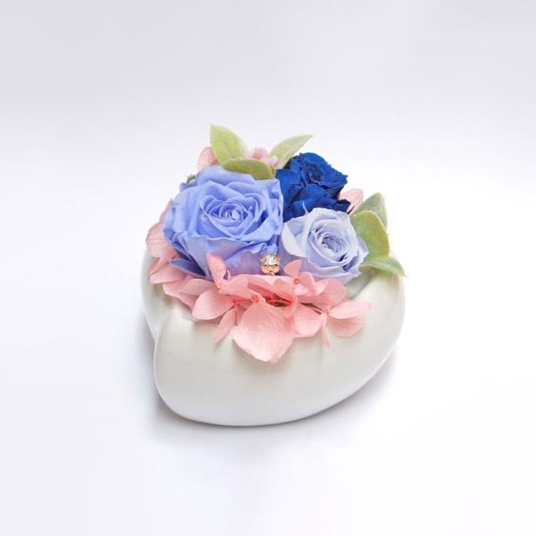 ハート型の丸いフォルムの陶器の花器に、爽やかな青色のプリザーブドフラワーが咲くアレンジです。お誕生日のプレゼントやお礼の品など、様々な用途にご利用いただけます。主な花材：薔薇、アジサイ（以上、プリザーブドフラワー）、リーフ（造花）、ガラスピ...