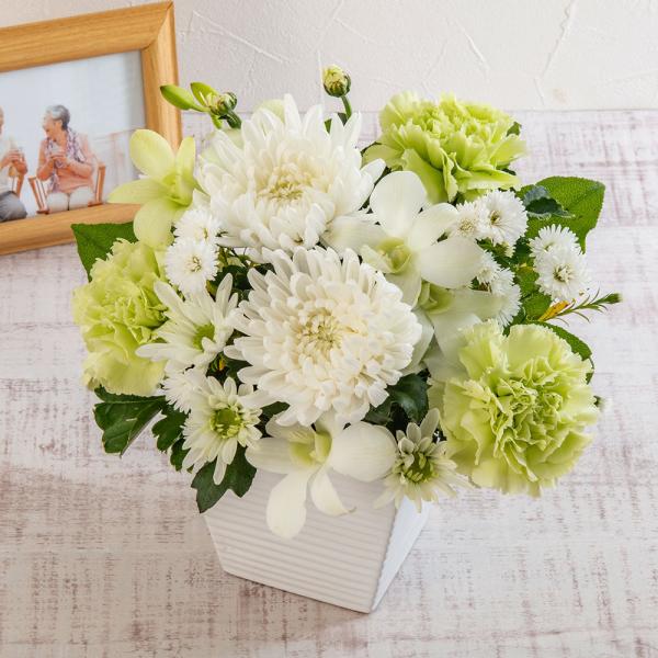 ■お届け 3日後以降でご指定可■注文締切2026/10/28　14:00まで清らかに咲く白い大輪のマムを中心に、カーネーションやラン、グリーンを白い陶器にアレンジしました。白地の花器を柔らかに取り囲む平衡の線が美しいデザインは、見る方の心が...