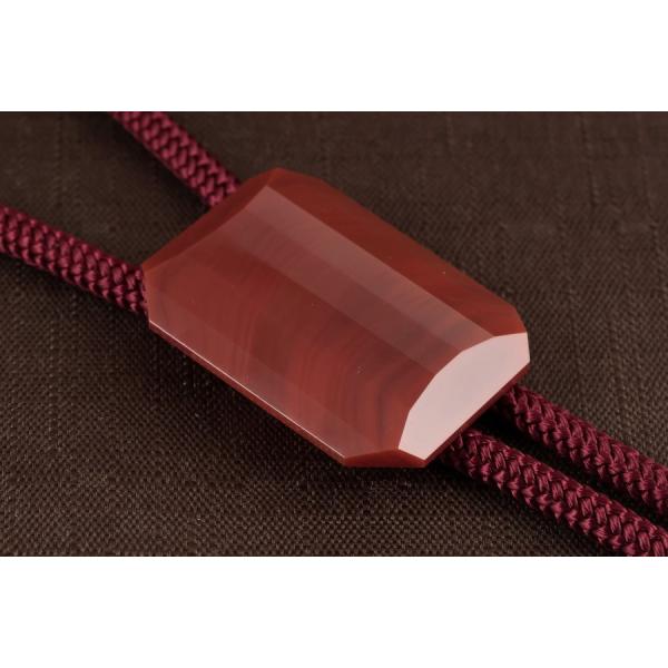 天然石で作ったループタイです。石の縁に施されたカットが、光を受けるとキラリと反射します。フレームがないため、石そのものの模様と質感を感じることのできるデザインです。朱色の石に薄っすらと縞模様が入ったメノウを使用しています。光に透かすと半透明...