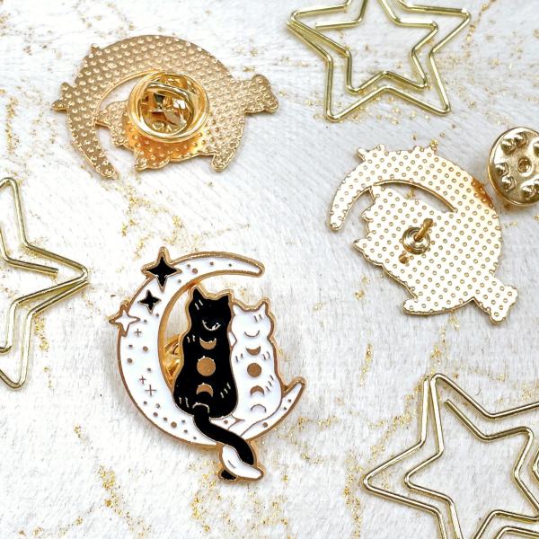 月で寄り添う２匹の猫をゴールドで縁取ったピンバッジです。猫の背中には『月の満ち欠け』がデザインされています。ブローチとして衣類に付けたり、後ろの留め具を外せば画鋲としても最適です。鞄や帽子などにもワンポイントとしてアクセントになります。【内...