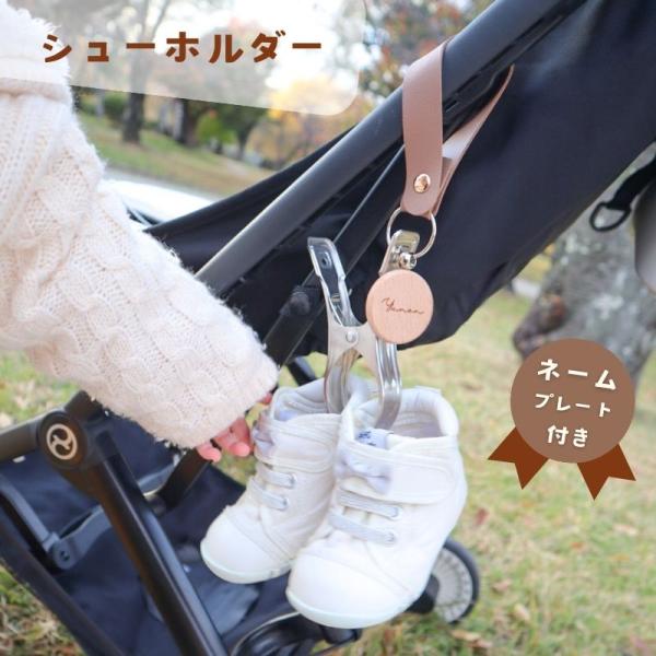 《　シューズホルダー　》ラテカラーのレザーバンドにゴールドのスナップボタンが付いたおしゃれなシューホルダーです。ネームプレートとして木製の丸プレートが付きます。赤ちゃんの靴だけでなく、帽子やおもちゃなどをベビーカーに引っ掛けておける便利アイ...