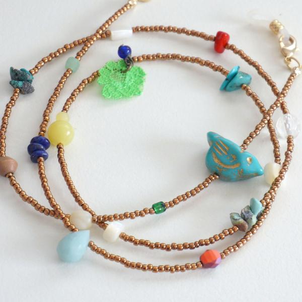 ブロンズのシードビーズをベースに天然石のターコイズやシェル、キラキラのガラスビーズなどが飾られたメガネチェーンです。可愛いターコイズ色の鳥のビーズとグリーンのレースがポイントです。【サイズ】全長約 76cm【素材】ビーズ、天然石、レース、真...
