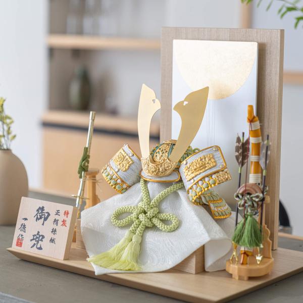 仕上の丁寧さに加え、個性的な素材を使った鎧兜が魅力の一徳作の兜。 兜はコンパクトながら純金箔貼の鋳物で花模様があしらわれ高級感があります。兜の顎紐(忍び緒)は若草色、兜の裾部分(錣(しころ))の糸は白で全体的に柔らかな印象の兜です。飾り台は...