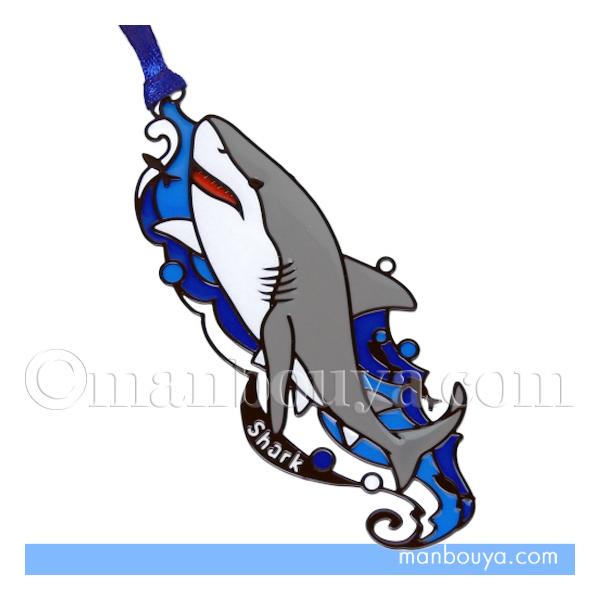 サメデザインのステンレス製ブックマーカー。本などに挟んで目印にする、しおりです。波と大柄のサメがダイナミックなデザイン。ブルーの部分とサメの口は半透明のステンドグラスタイプになっています。裏面は鮫が黒になっていて、赤い口だけが見えるシルエッ...