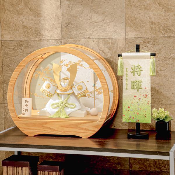 白木にアクリルケースのため軽い設計となっています。兜は黒いのが多い中、白地に金で縅の糸は若草色と明るく五月に相応しいお飾りです。四角いケースが多い中、丸いケースは珍しく長めに飾ってあげてください。間34×奥24×高30