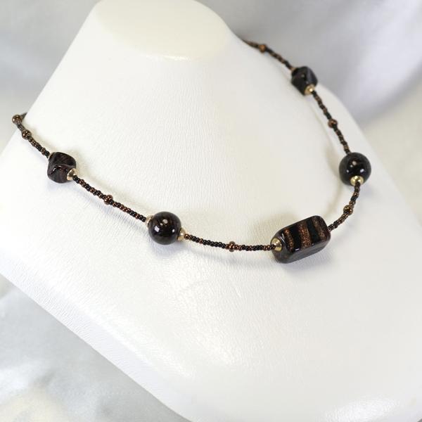 角柱ビーズ、丸ビーズ、変形ビーズを左右対称に配したステーションネックレスです。黒ベースのビーズ入れた金茶の模様が上品に輝きます。メインのビーズを引き立てるように、ネックビーズにはメタリック系のブラウンと黒をランダムに使用。雰囲気のある、手の...