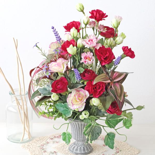 母の日に贈る、ミニバラとカーネーションのアレンジメントです。アンティーク調の器を使い、深みのある色合いのリーフを添えた赤い花々がシックな印象です。アレンジメントサイズ：約高さ36cm×幅27cm×奥行17cmシルクフラワー(造花)は枯れない...