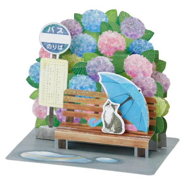 バス停のそばにアジサイが咲いている風景を立体的に表現しました。ベンチの上にいるネコ、誰かがさしてくれた傘、水たまりに映る虹などからストーリー性を感じられるカードです。ラメ加工が施されており、キラキラと輝いて雨上がりの雰囲気を出しています。立...